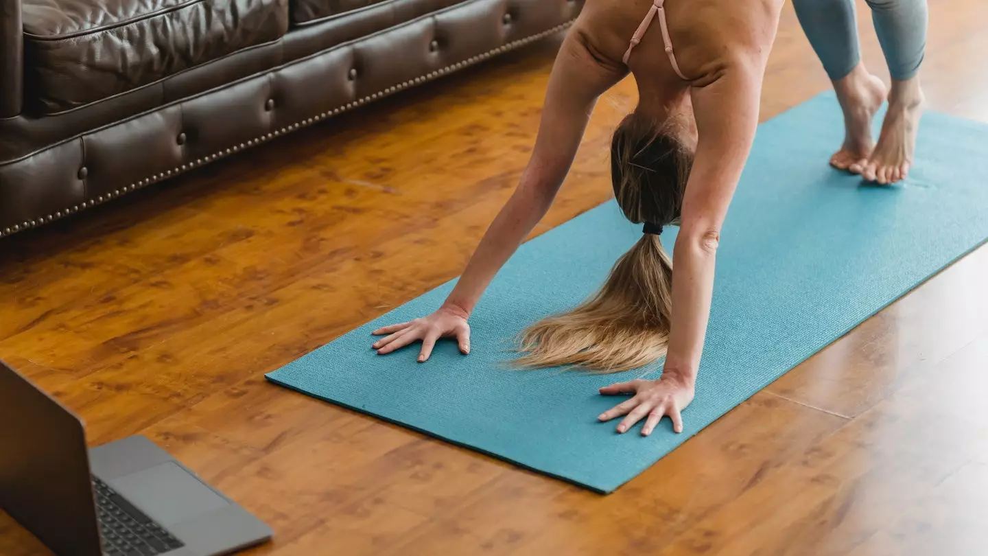 Uma mulher loira esticando ou fazendo ioga em um tapete de ioga em um piso de madeira na frente de um sofá com um laptop Macbook cinza na frente dela.